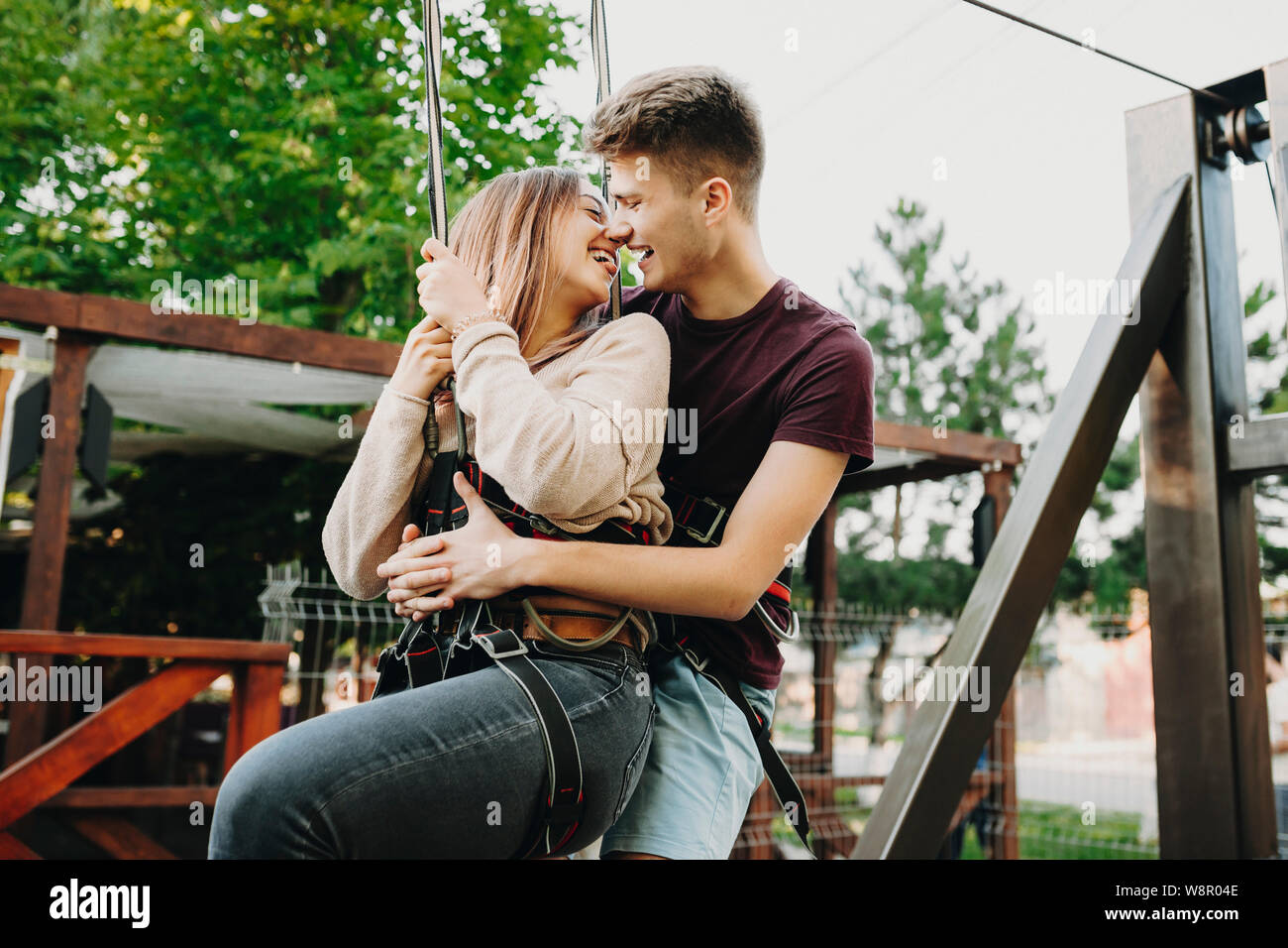 Embracing young man and woman in safety harness hanging on cable of zip ...