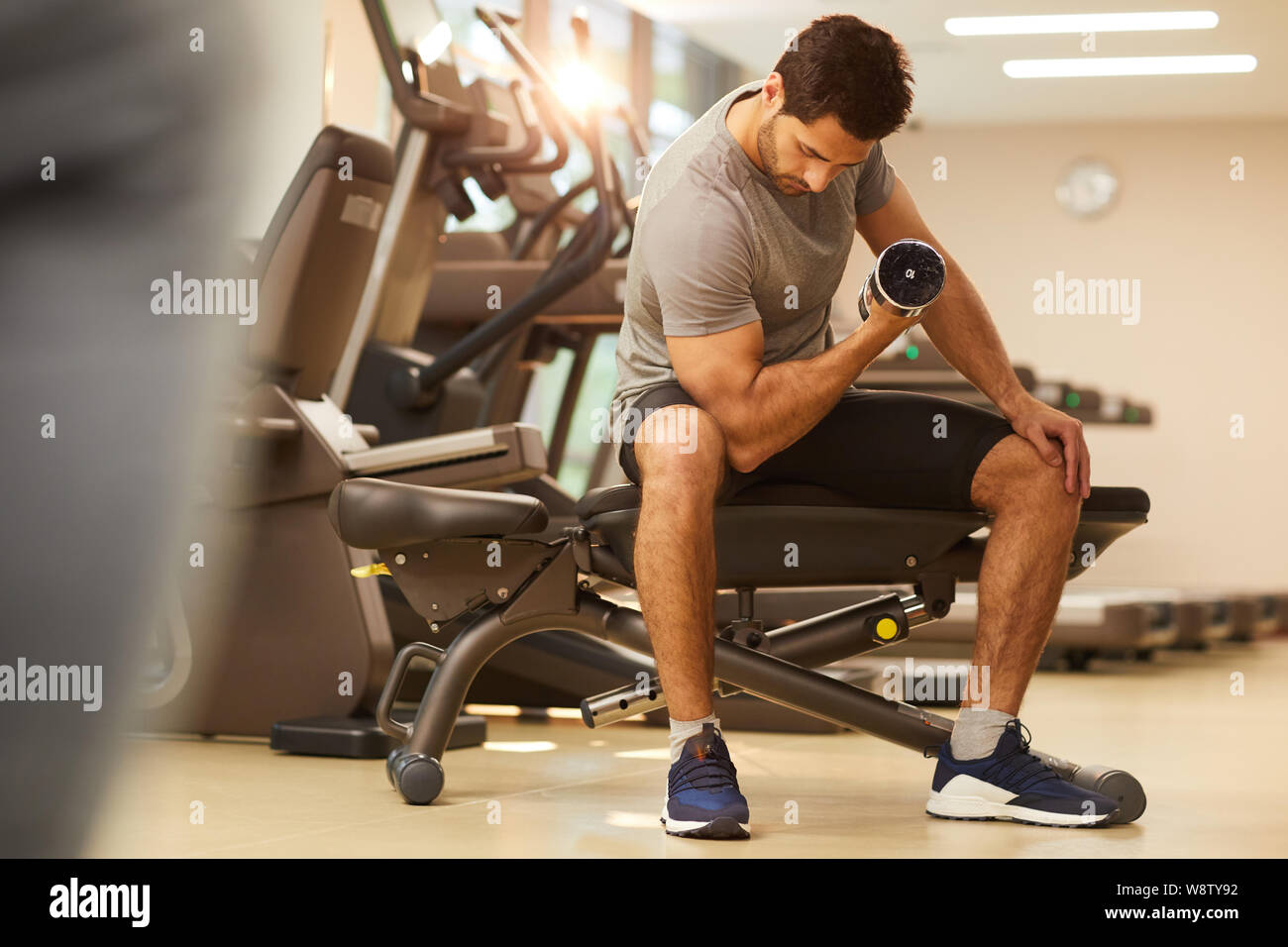 Full length portrait of muscular man lifting dumbbells sitting on bench ...