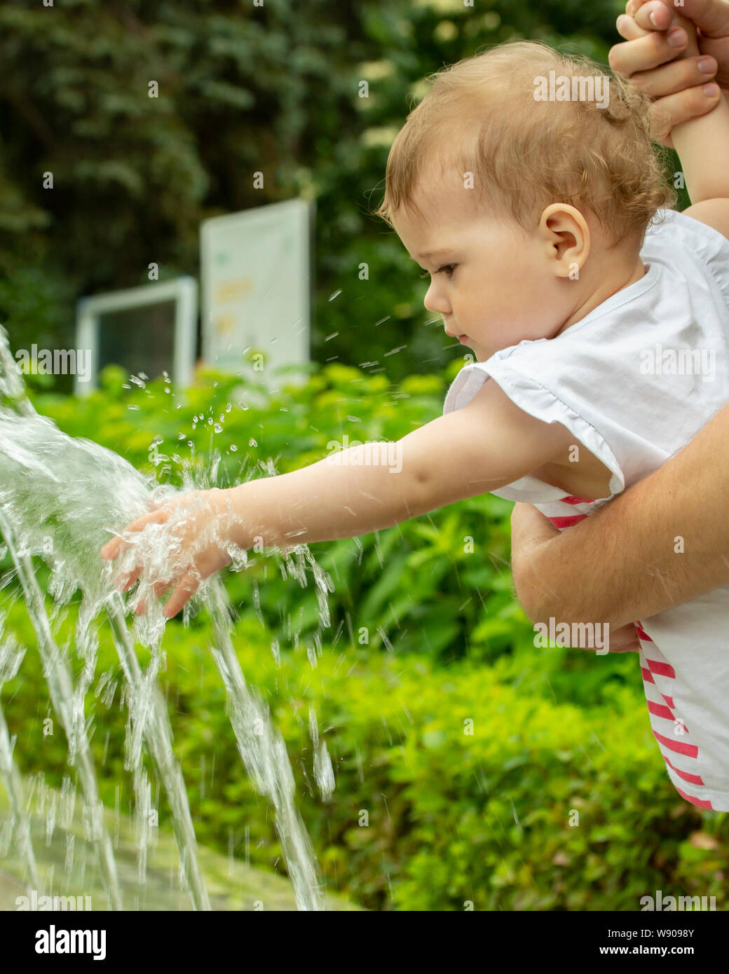 Baby girl stretches her hand to the fountain, the baby plays with jets ...