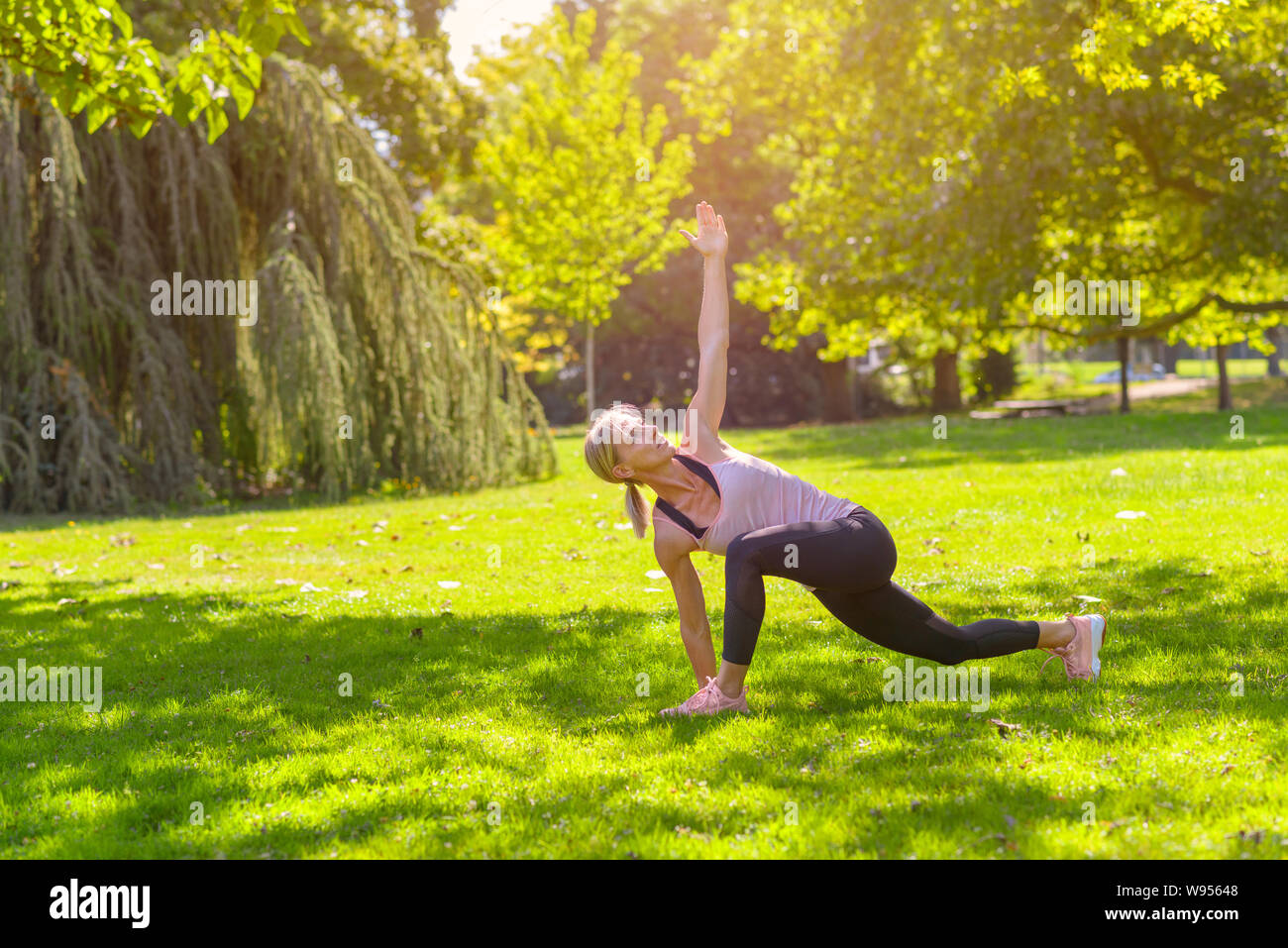 Fit woman doing lunge and stretch exercises outdoors on a lush green ...