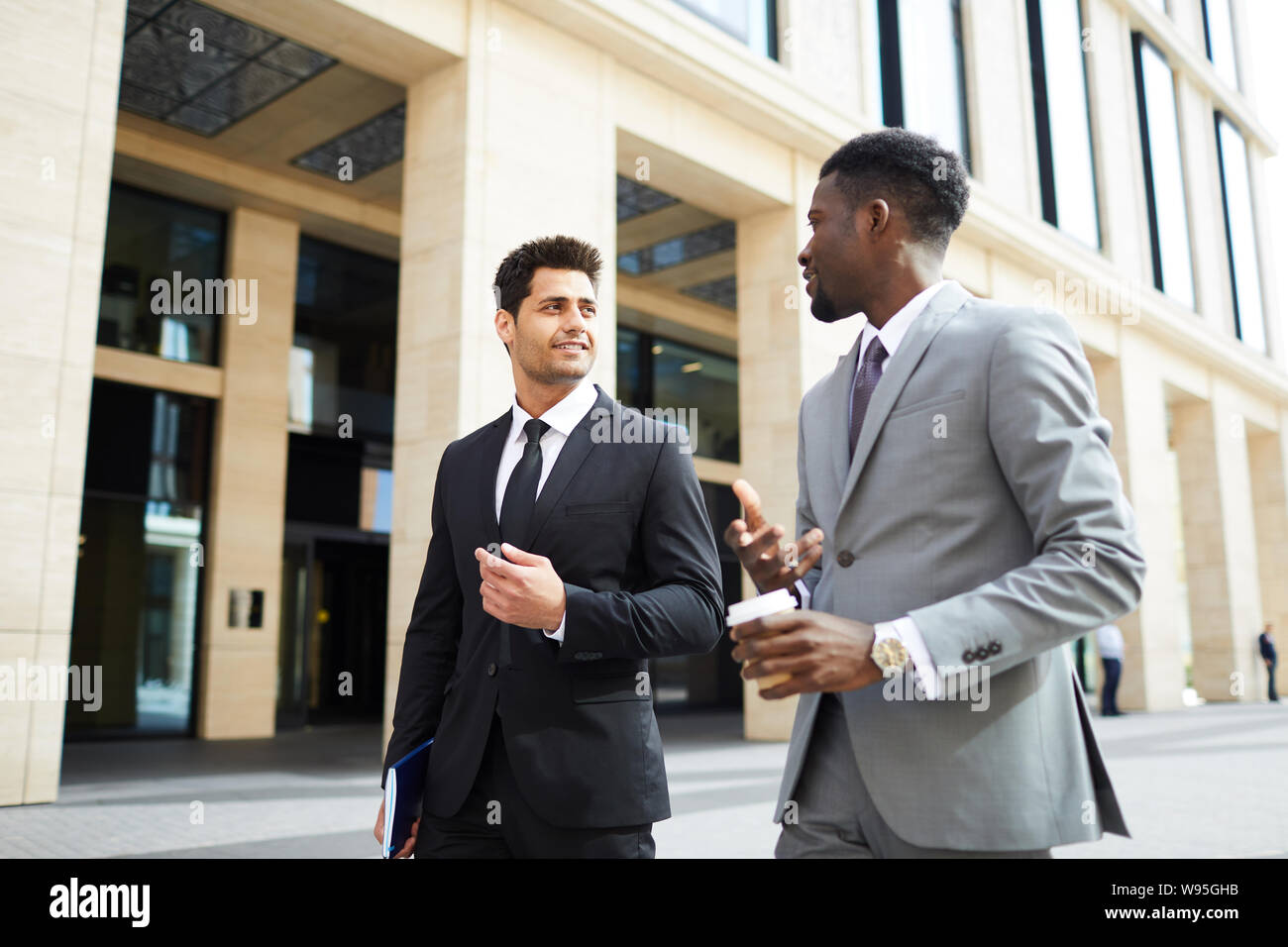 Two young businessmen in suits talking to each other while walking ...
