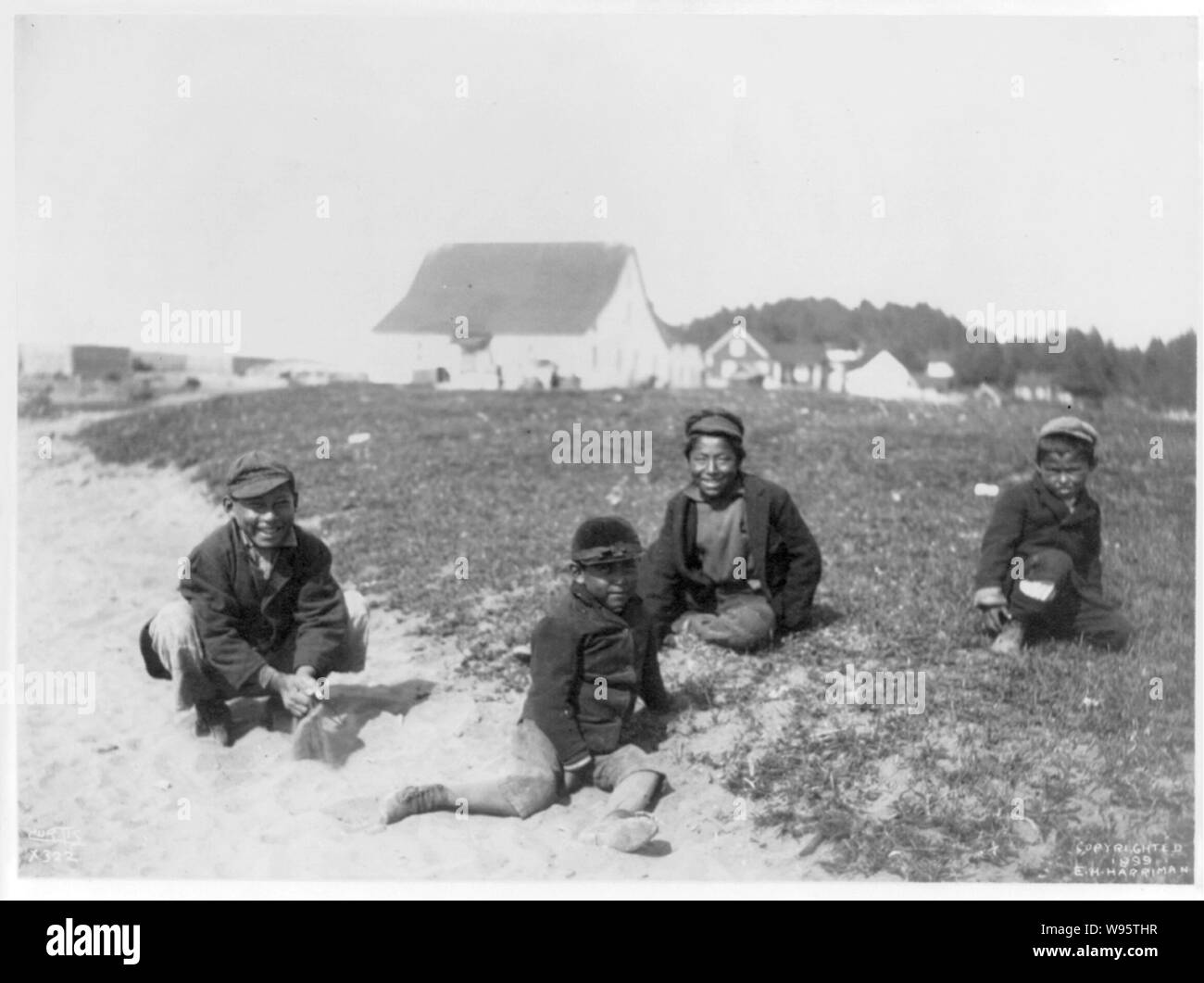 Aleutian Islands : 4 Indian boys seated on ground, buildings in backgrd ...