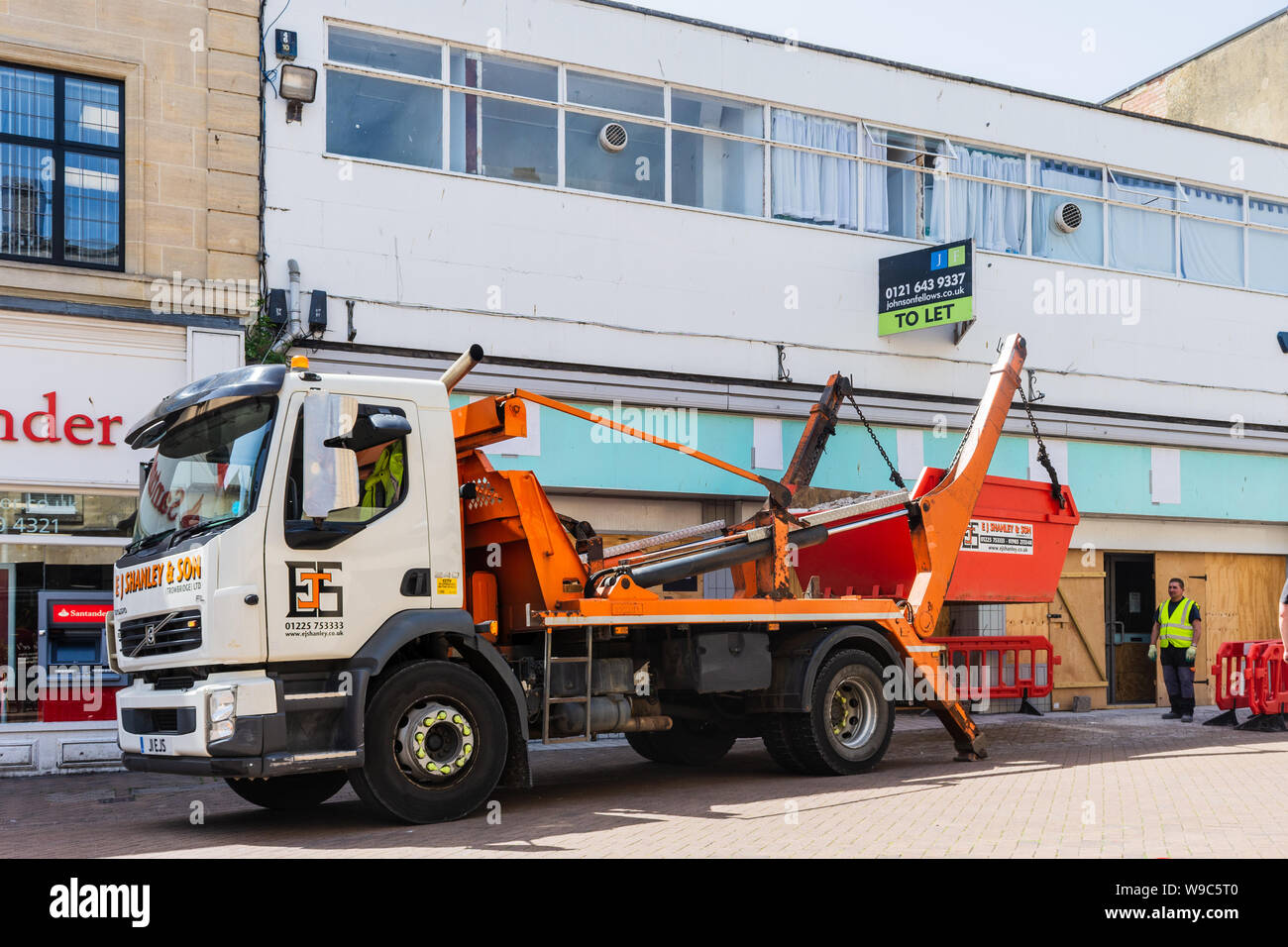 A skip lorry by a shop under refurbishment, with the driver using ...