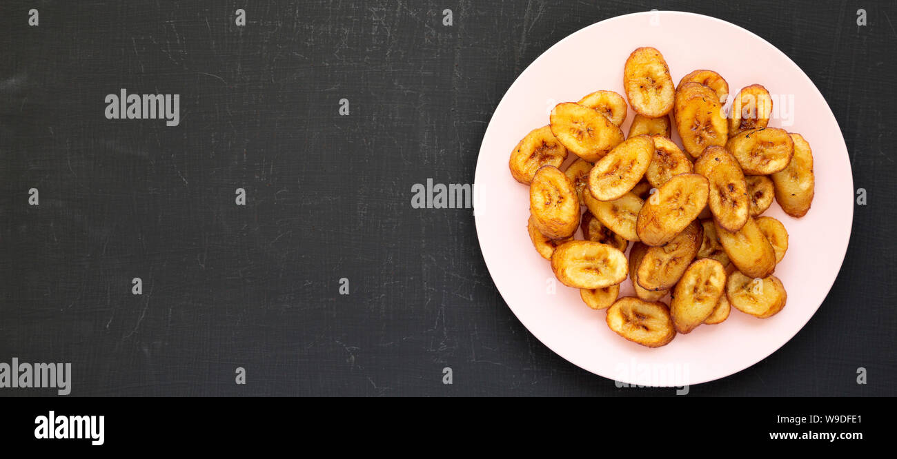 Homemade fried plantains on a pink plate on a black surface, overhead ...