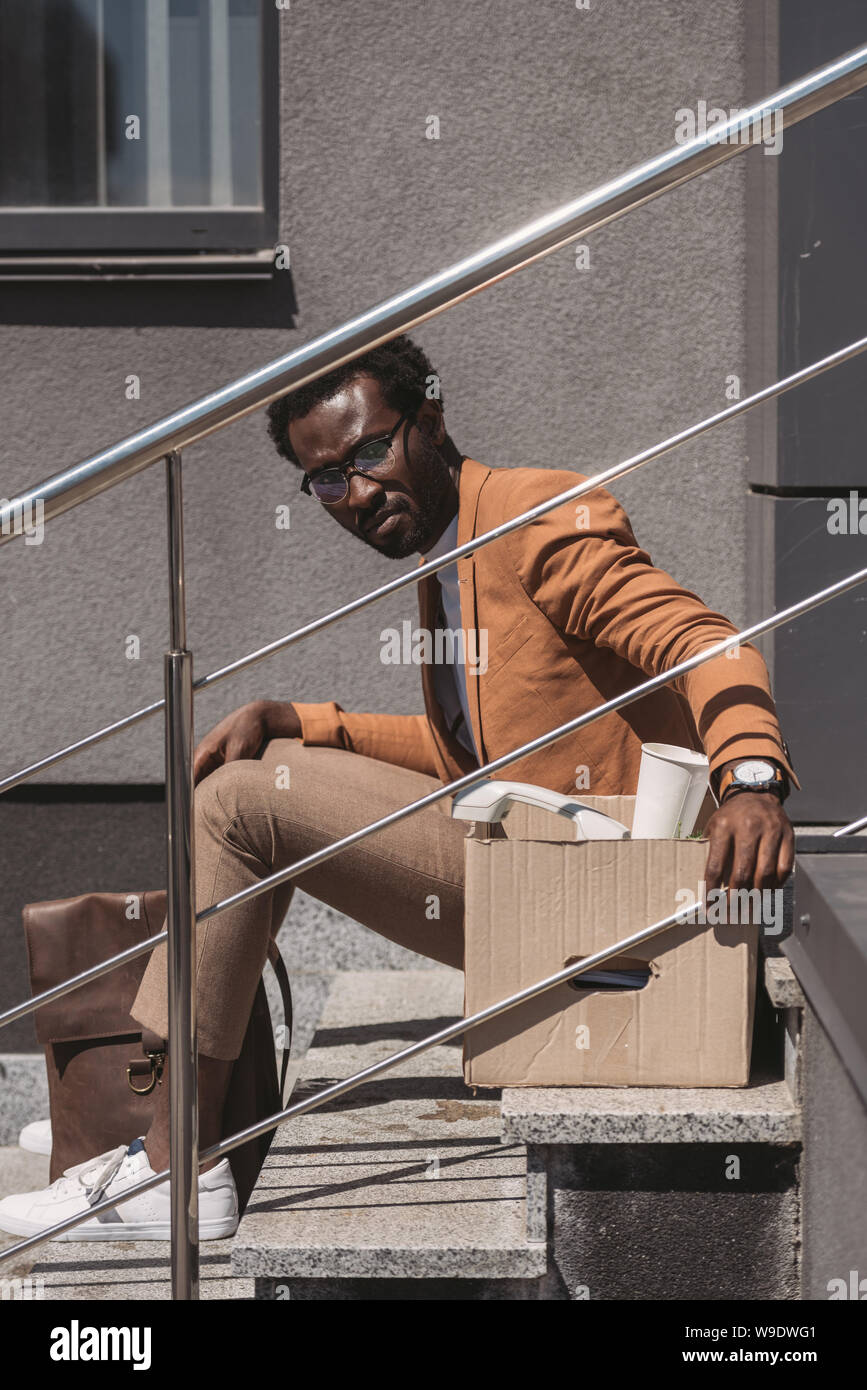 sad, dismissed businessman sitting on stairs on street near carton box ...