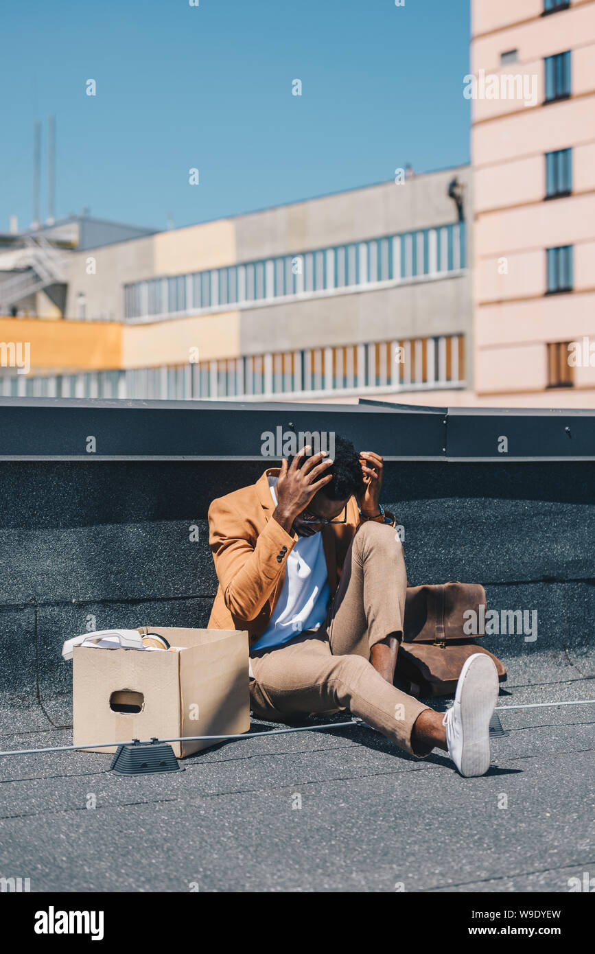 depressed african american businessman sitting on rooftop near carton ...