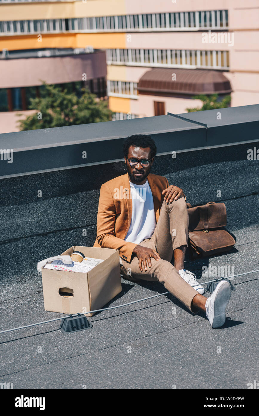 depressed african american businessman sitting on rooftop near carton ...
