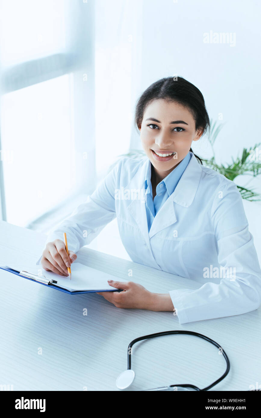 attractive latin doctor writing on clipboard while smiling at camera in ...