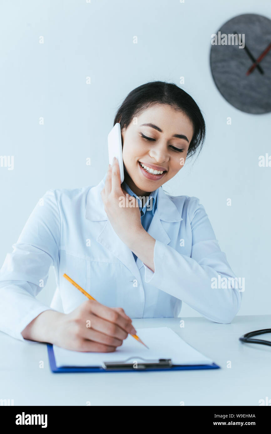 cheerful latin doctor writing diagnosis on clipboard while talking on ...