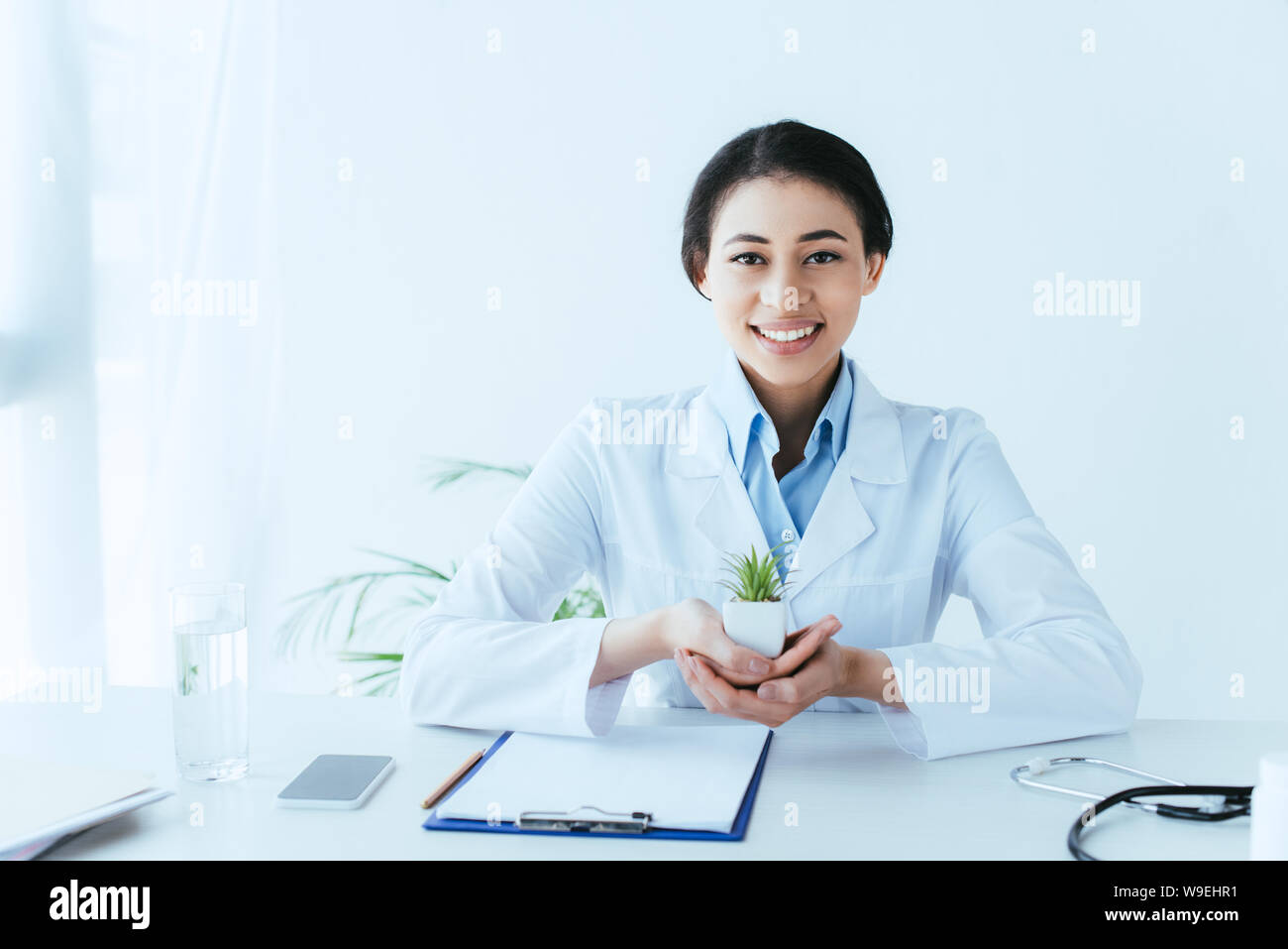 attractive latin doctor holding potted plant while sitting at workplace ...