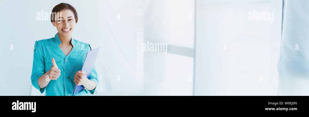panoramic shot of pretty latin doctor holding clipboard and showing ...