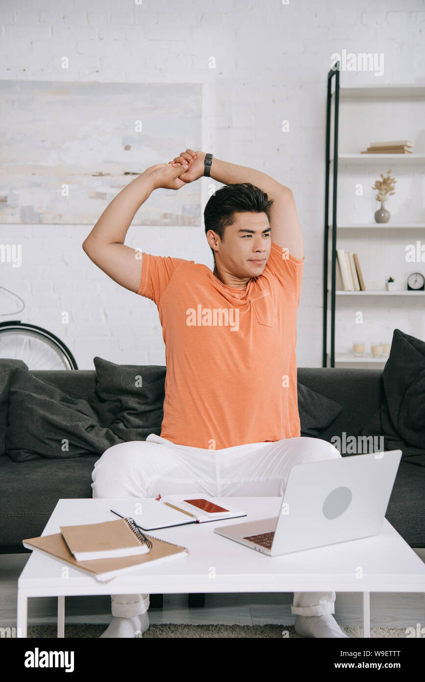 tired asian man stretching and looking away while sitting on sofa near ...