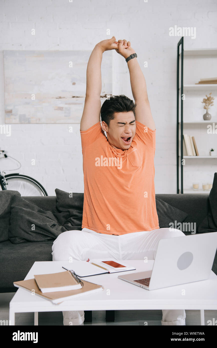 tired asian man stretching and yawning while sitting on sofa near table ...