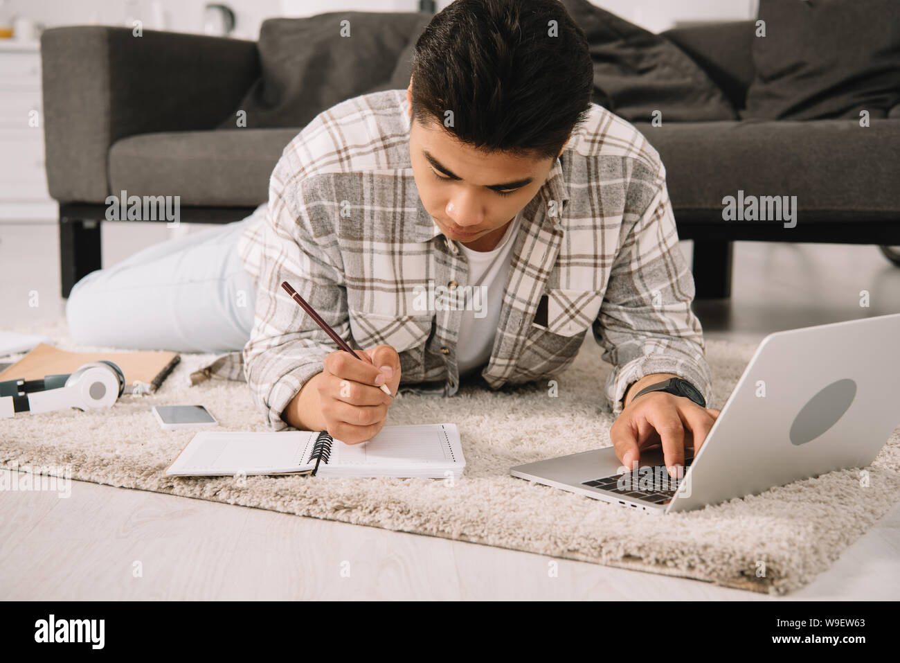 attentive asian man writing in notebook and using laptop while lying on ...