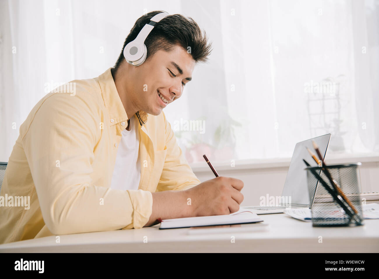 cheerful asian man in headphones writing in notebook while sitting at ...