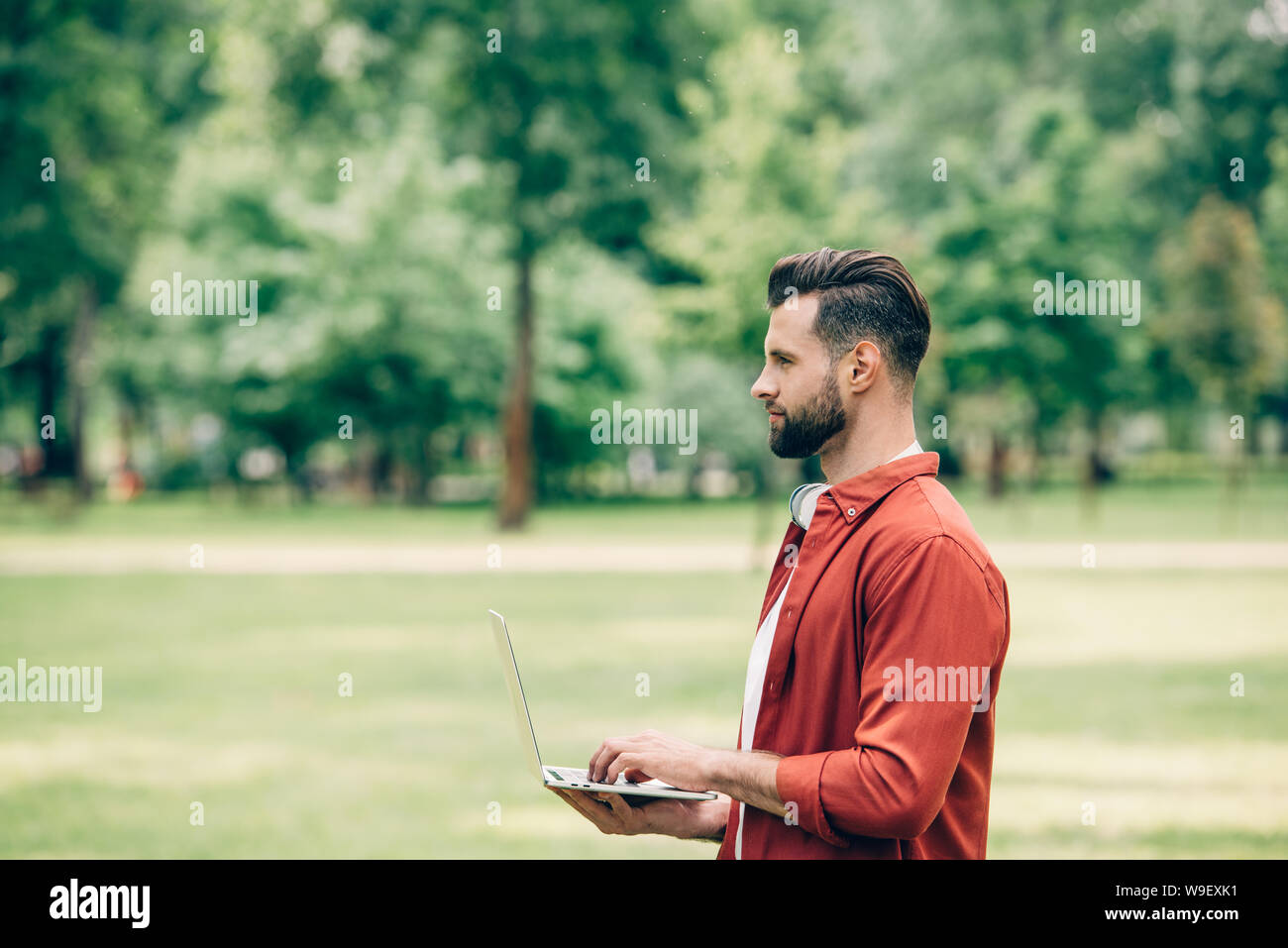 Side view of young man standing in park with laptop in hands and ...