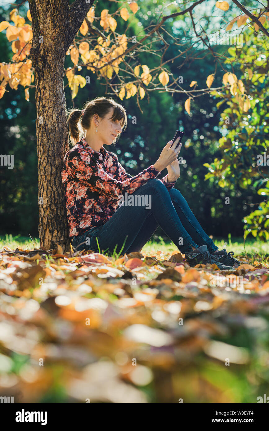 Young woman sitting under an autumn tree in jeans and jacket browsing ...