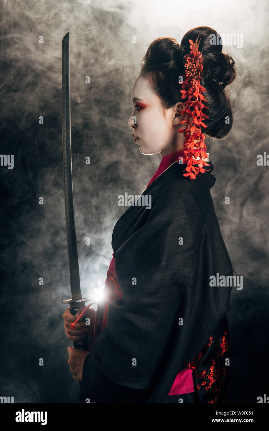 side view of geisha in black kimono holding katana in smoke on black ...
