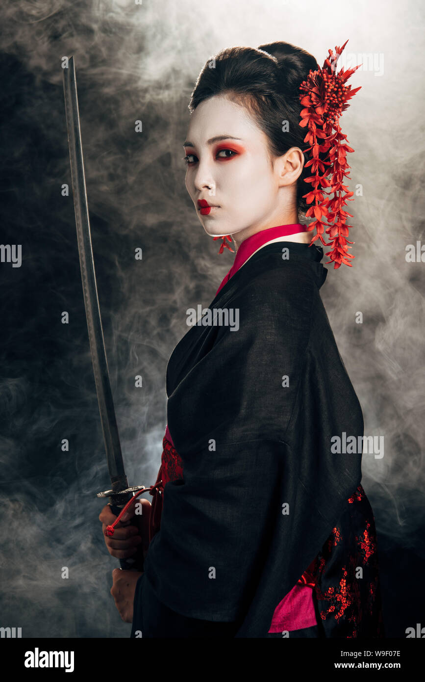 side view of beautiful geisha in black kimono holding katana in smoke ...