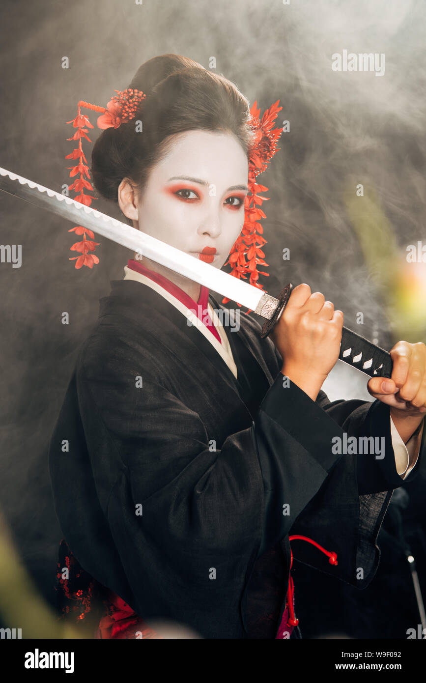 selective focus of geisha in black kimono holding katana in smoke and ...