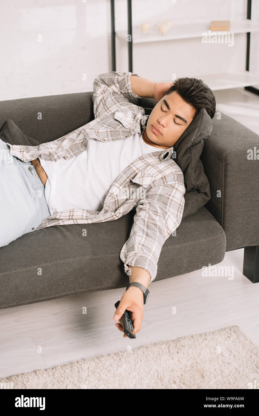 exhausted asian man sleeping on couch while holding tv remote ...
