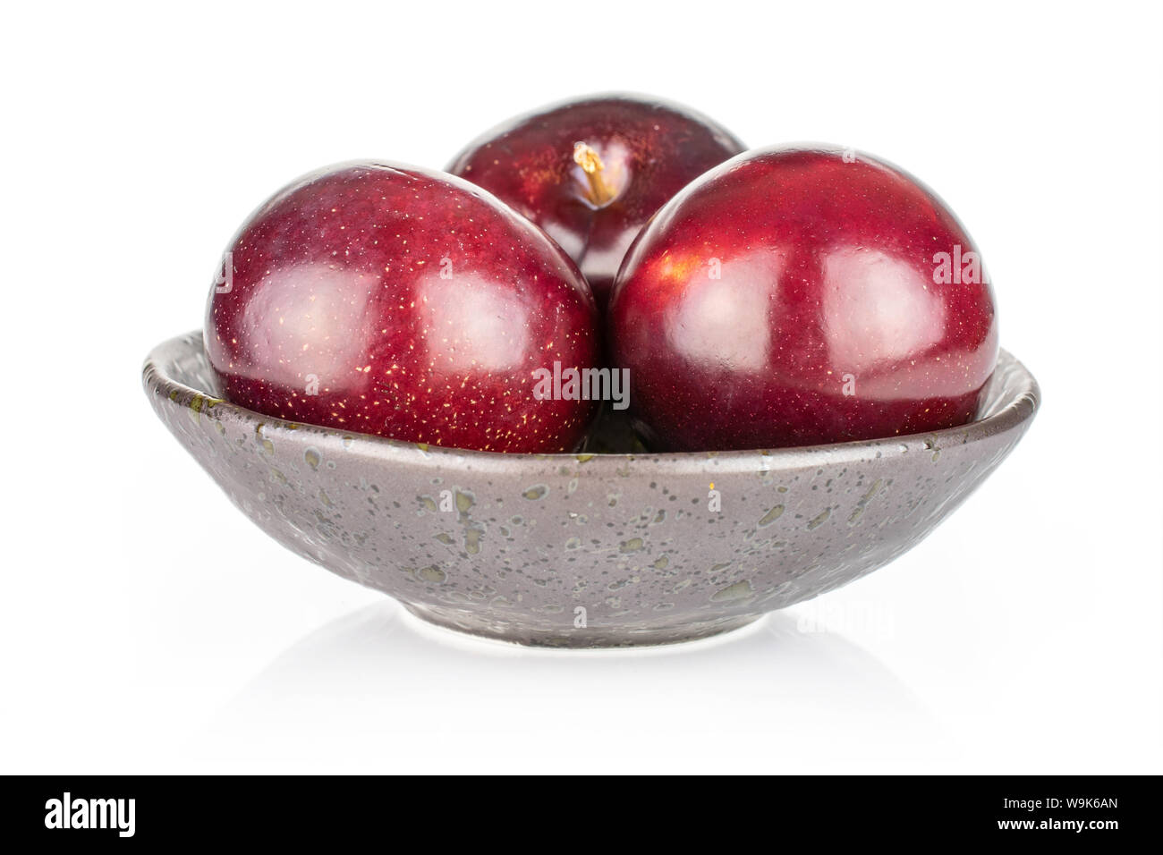 Group of three whole ripe red round plum in dark ceramic bowl isolated ...