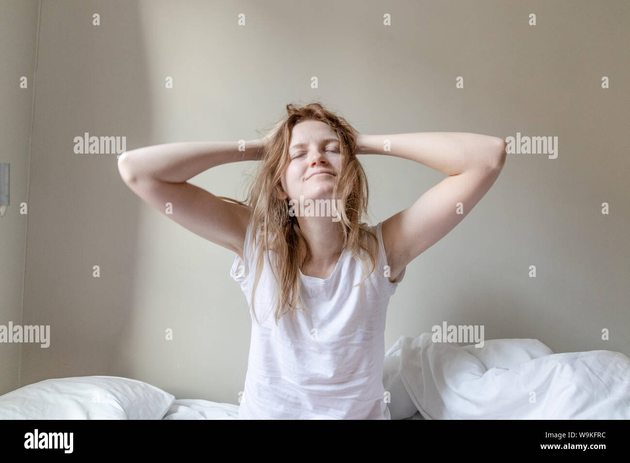 happy beautiful young woman waking up in morning, sitting on bed ...