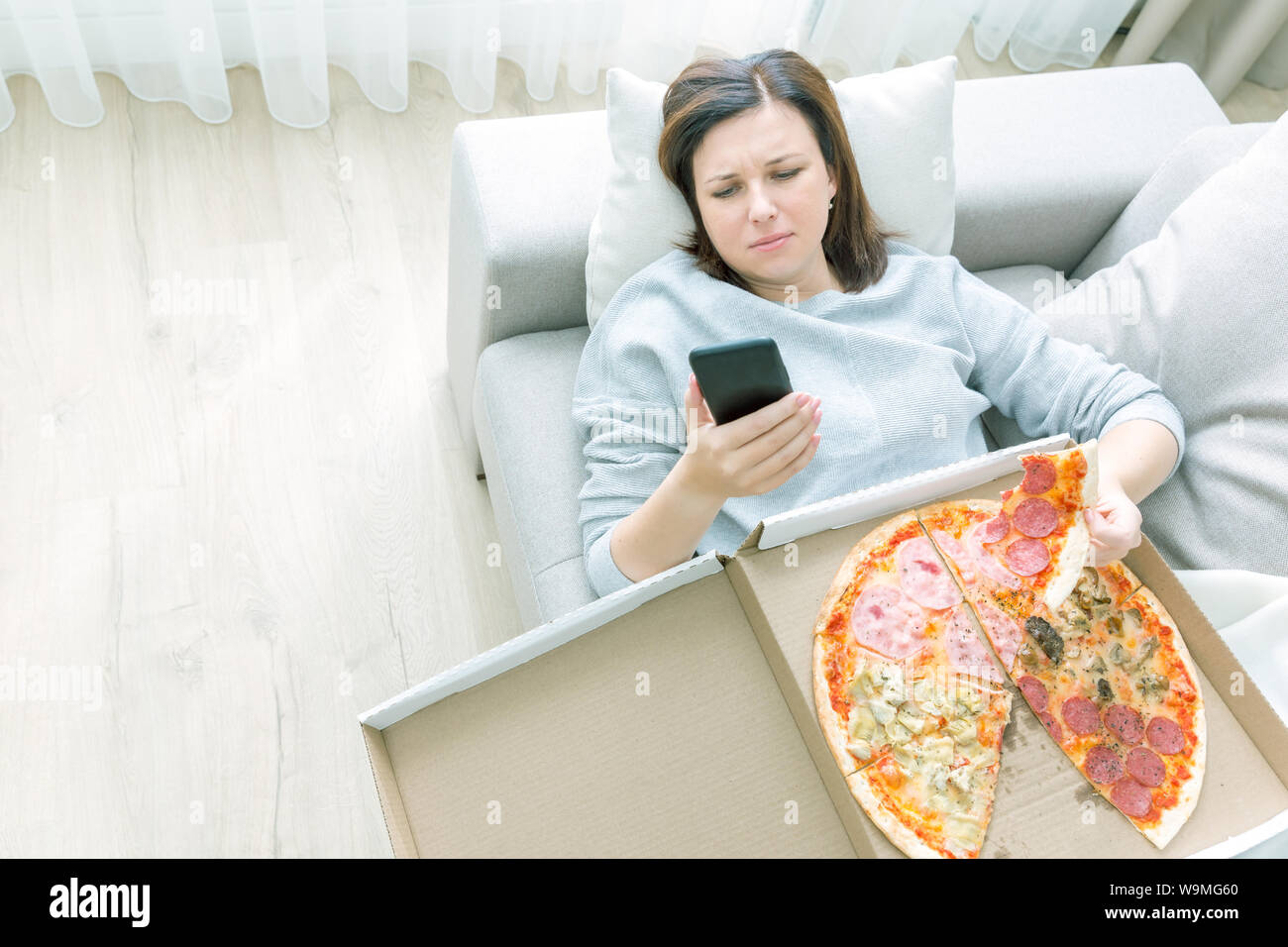 Sad woman eating pizza and holding phone laying on sofa at home, blue ...
