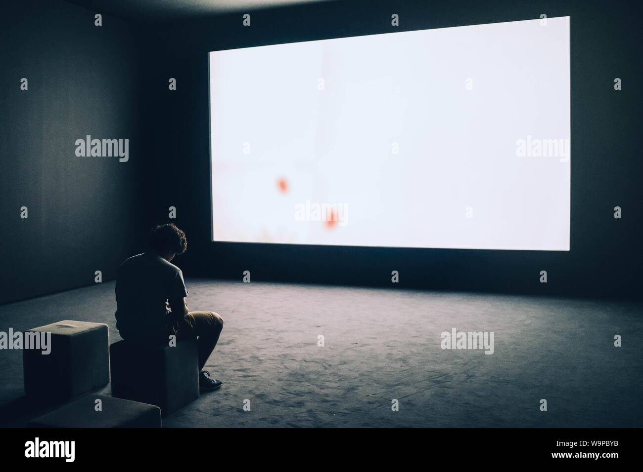 Artistic shot of a man sitting in front of a large white screen with ...