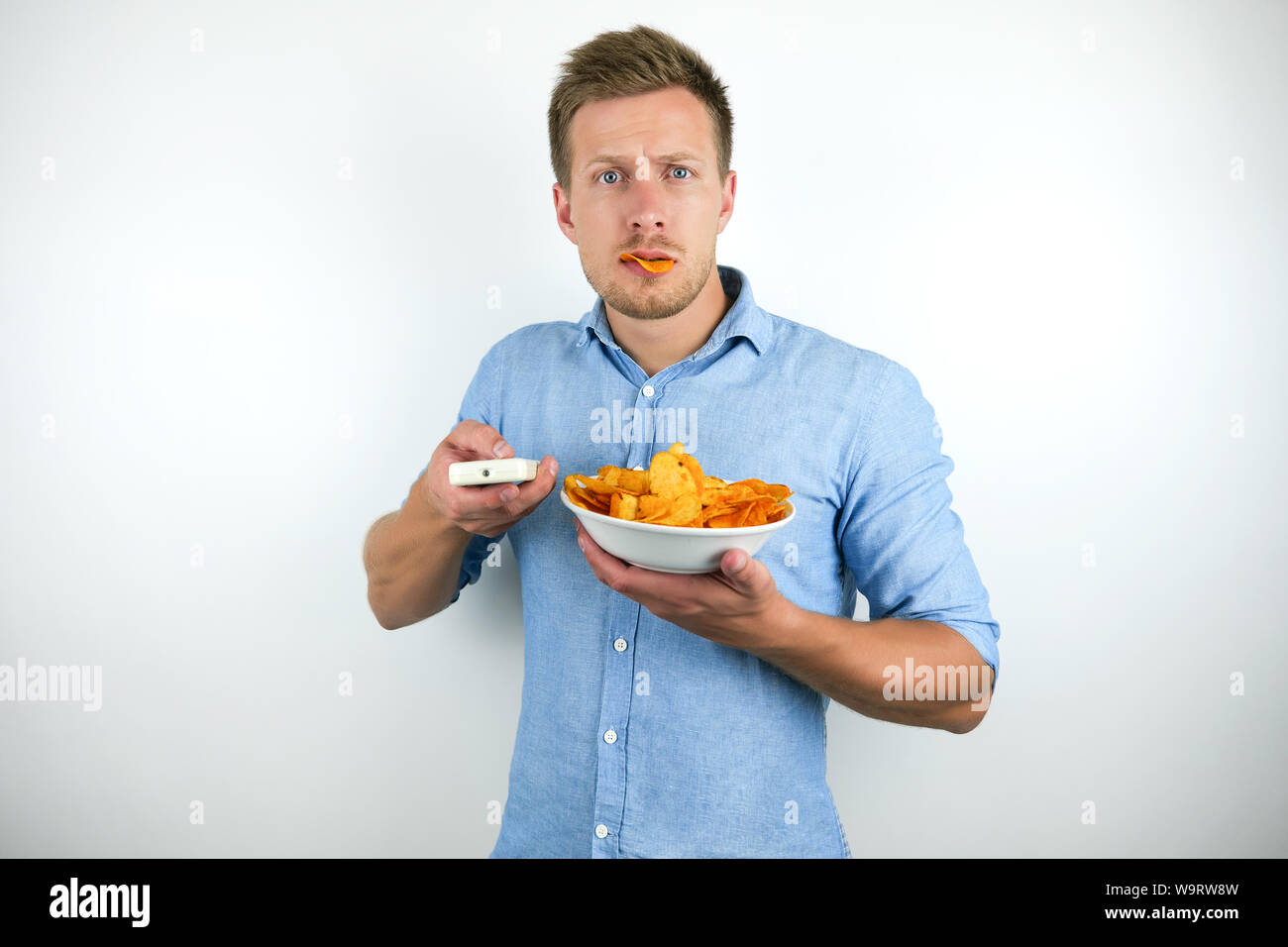 young handsome man eats chips holds remote control on isolated white ...