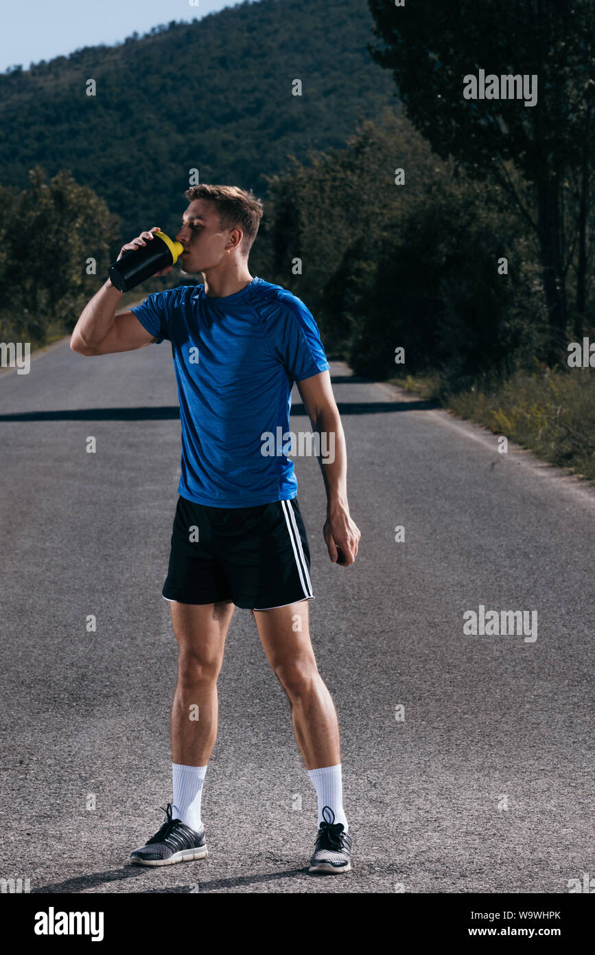 Fit male runner drinking water from a water bottle on an empty road in ...