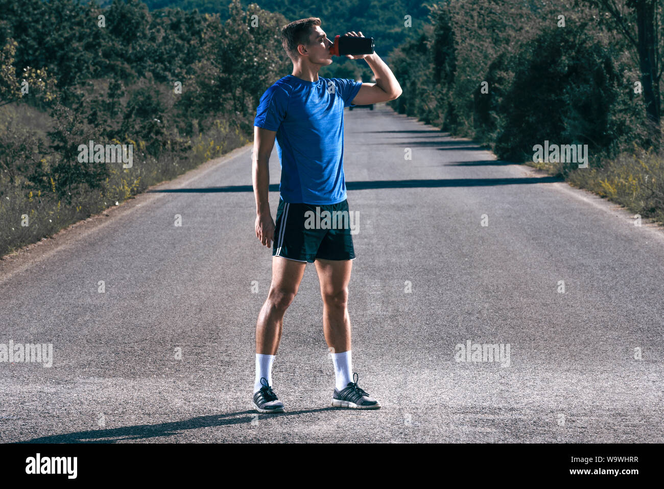 Fit male runner drinking water from a water bottle on an empty road in ...