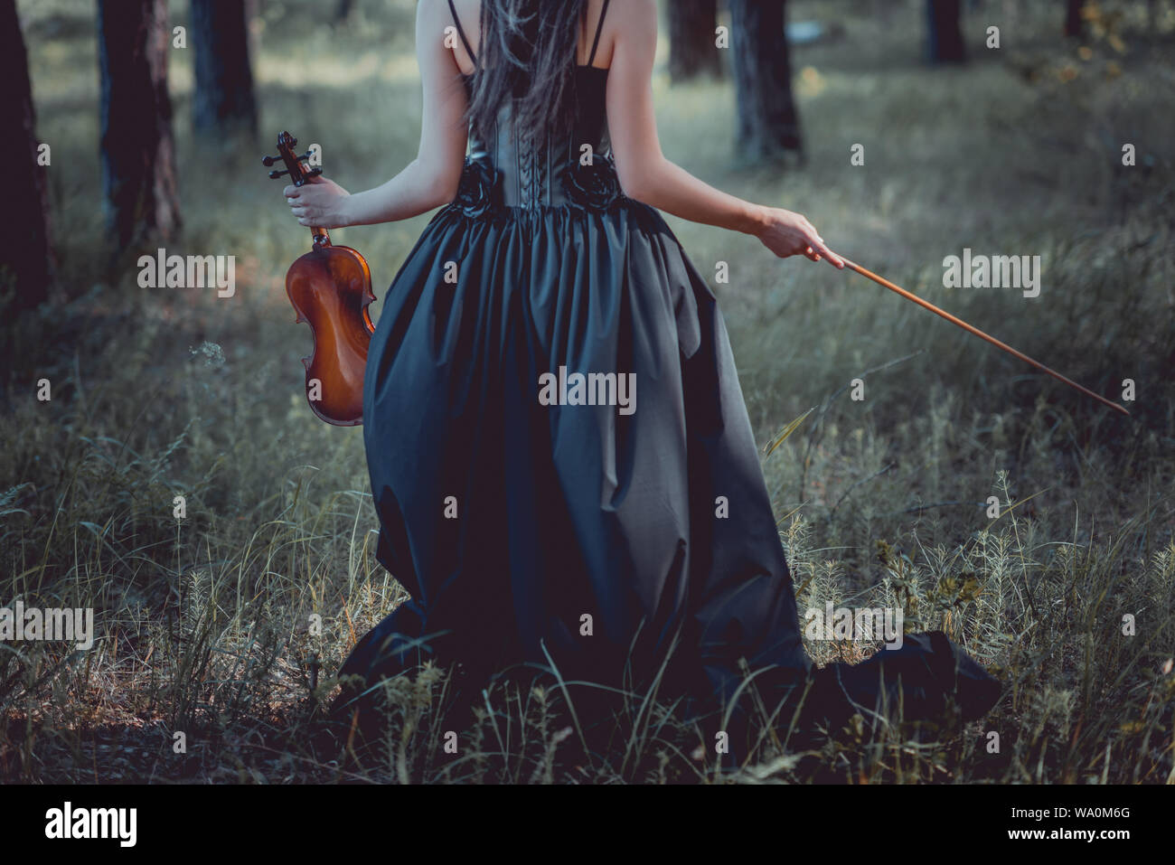 back view of woman in witch costume walking on forest, holding violin ...