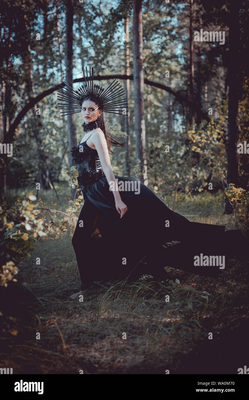 side view of brunette woman in witch costume standing on forest ...