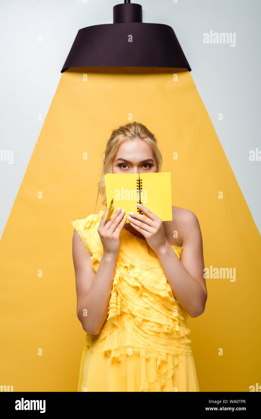 blonde young woman covering face with notebook on white and yellow ...