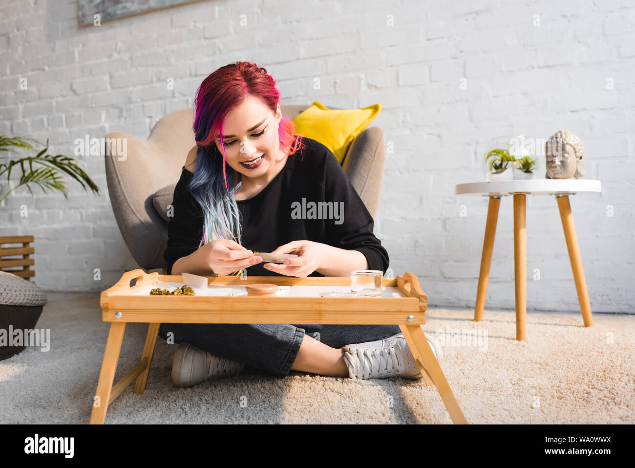 attractive hipster girl sitting on floor behind table and rolling joint ...
