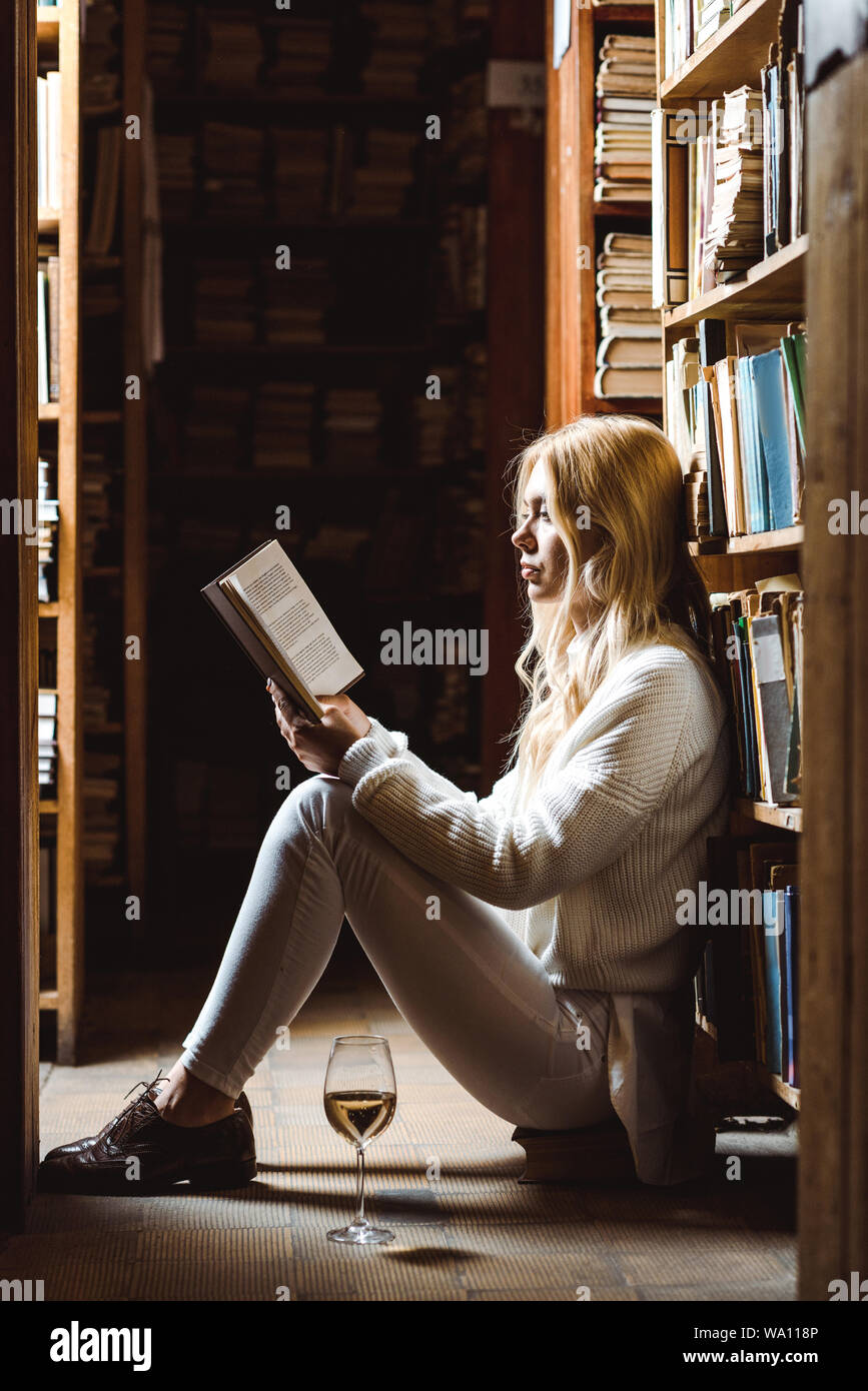 side view of blonde woman reading book and sitting on floor in library ...