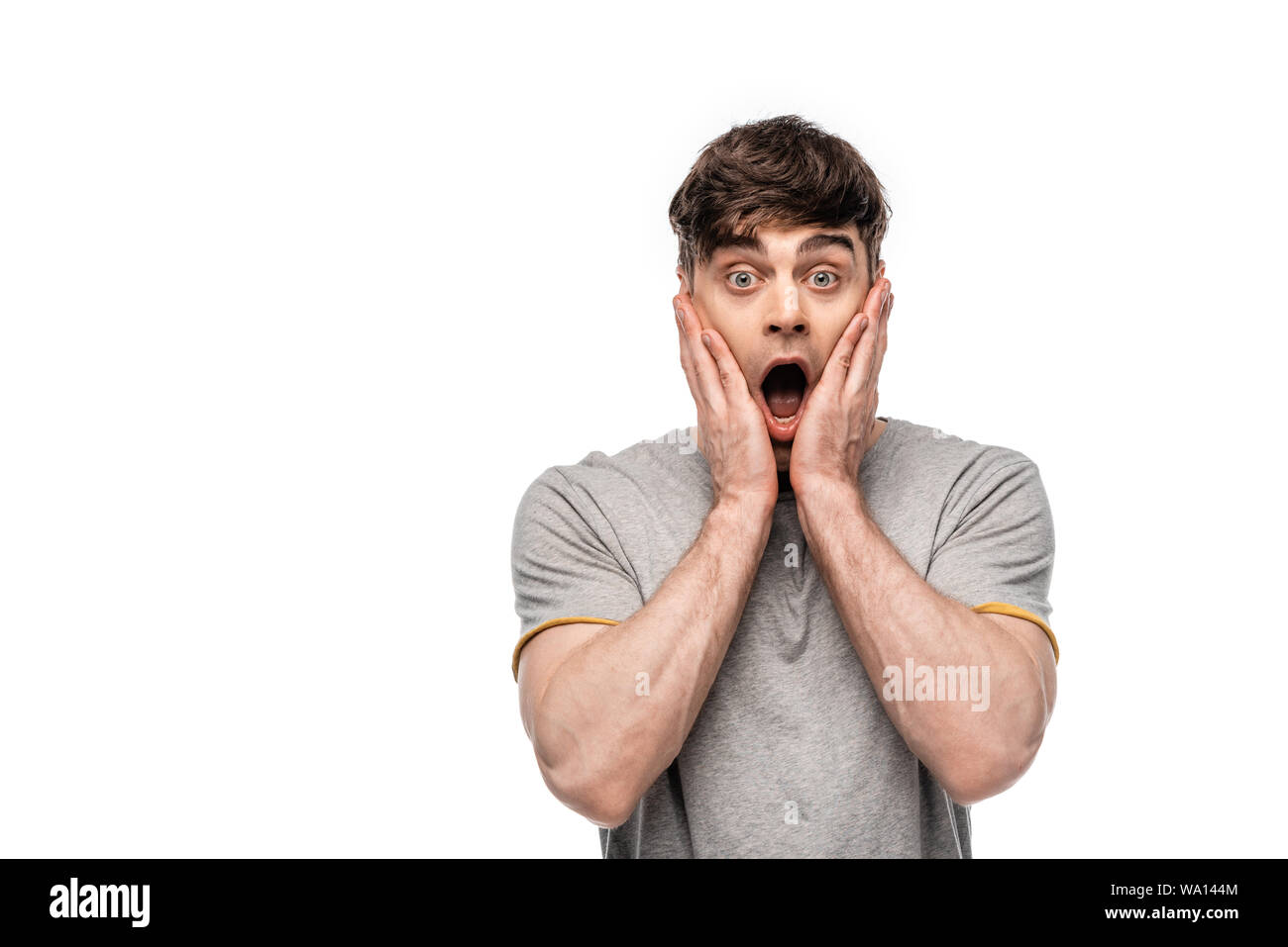 shocked young man looking at camera while holding hands near face ...