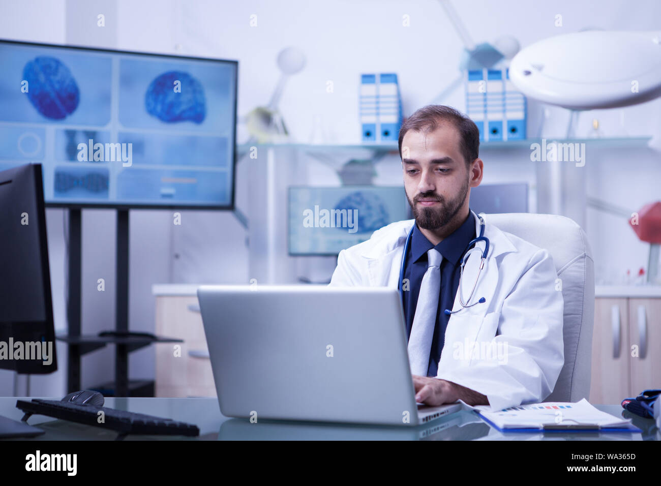 Doctor working on his laptop sitting at his desk in the hospital ...