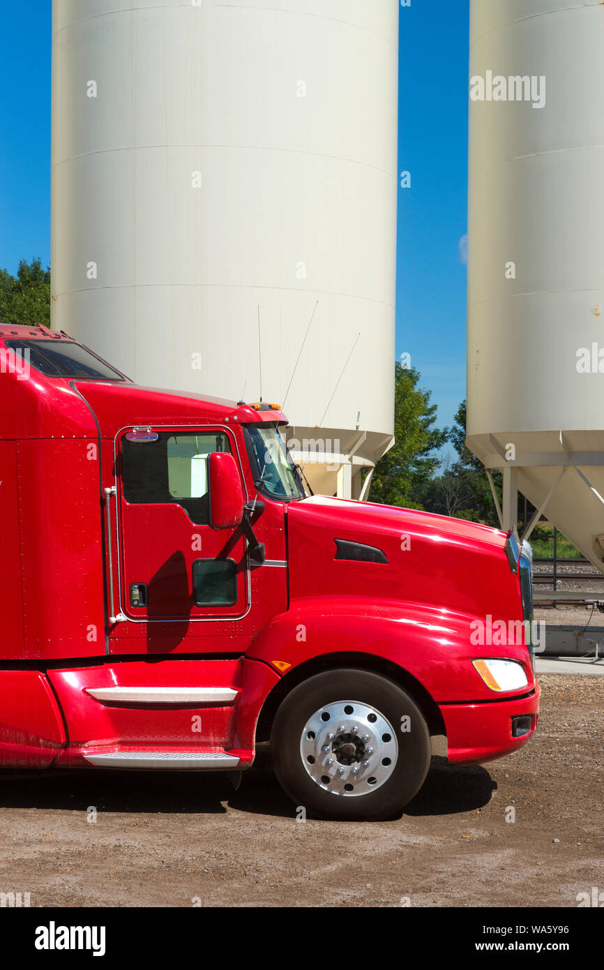 A red and black semi truck trailer parked alongside white grain storage ...