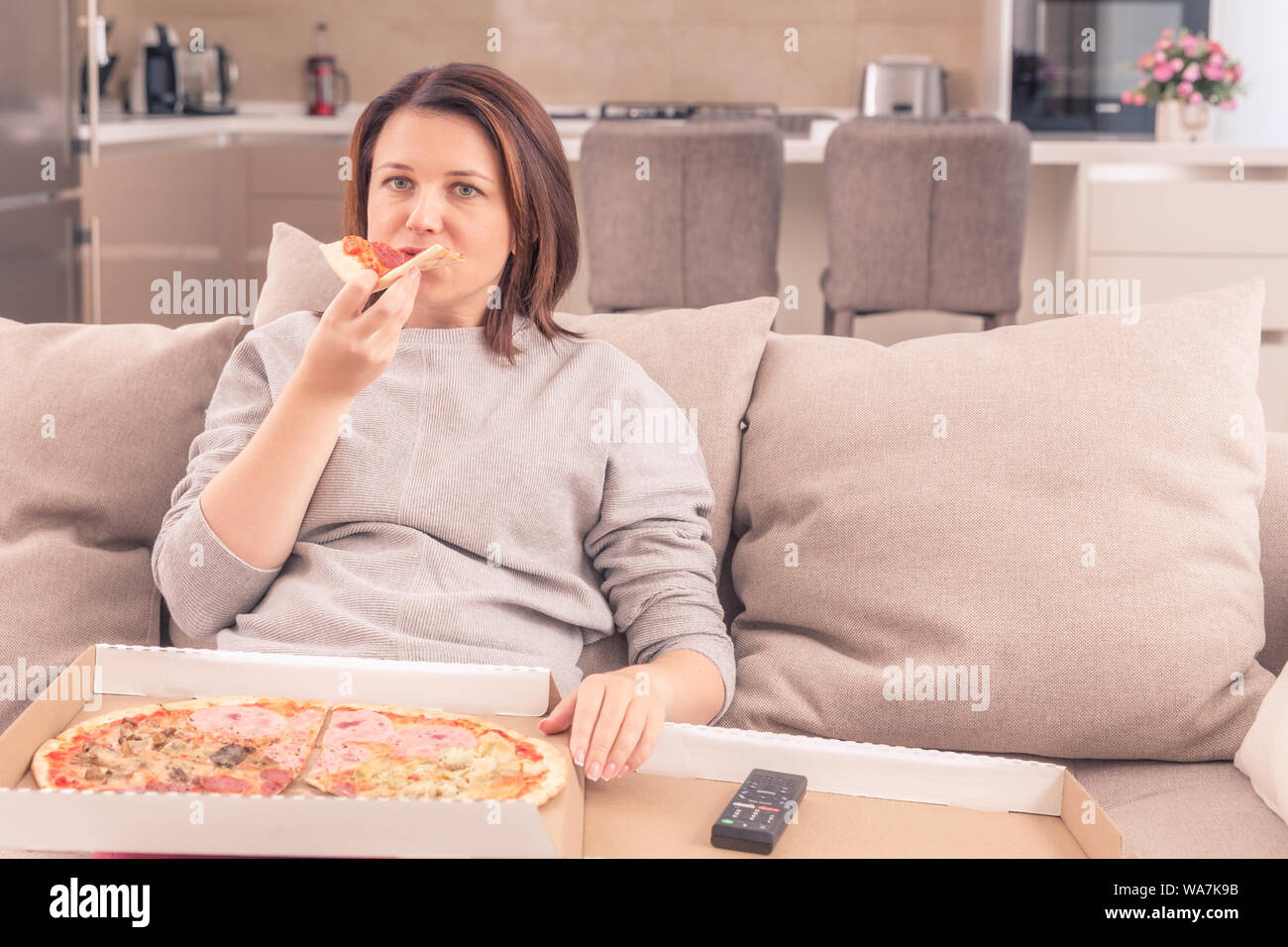 Surprised woman eating pizza and watching TV with remote control at ...