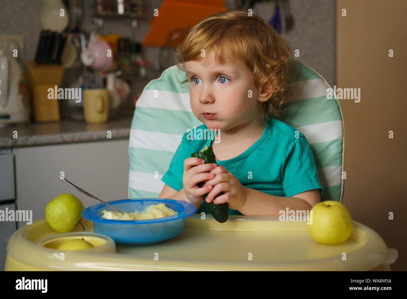 Charming little child eating in the kitchen. He sits in a highchair ...