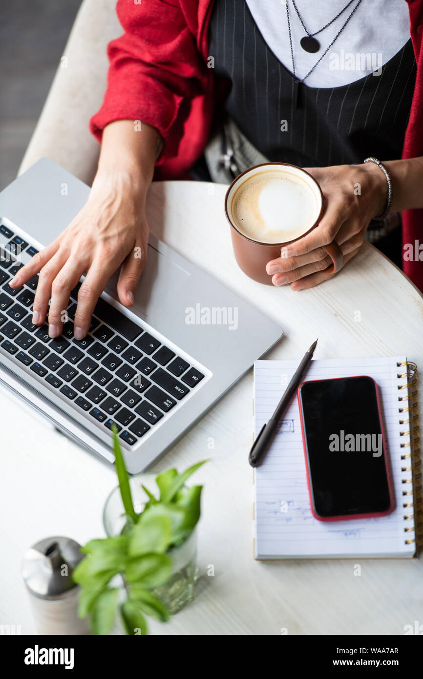 Young woman with cappuccino pressing keys of laptop keypad by workplace ...