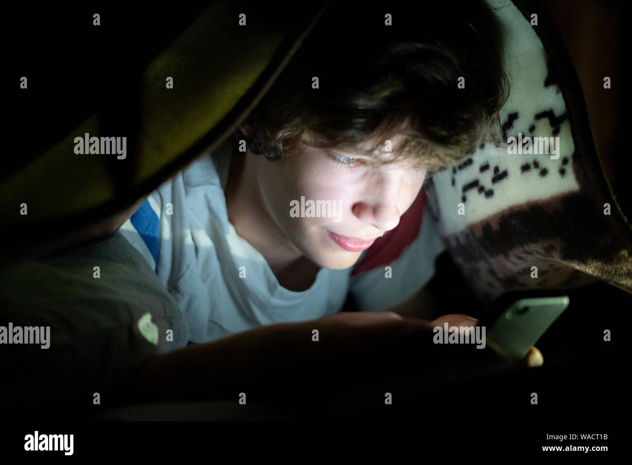 young boy using a smartphone lying in bed under a blanket at night ...