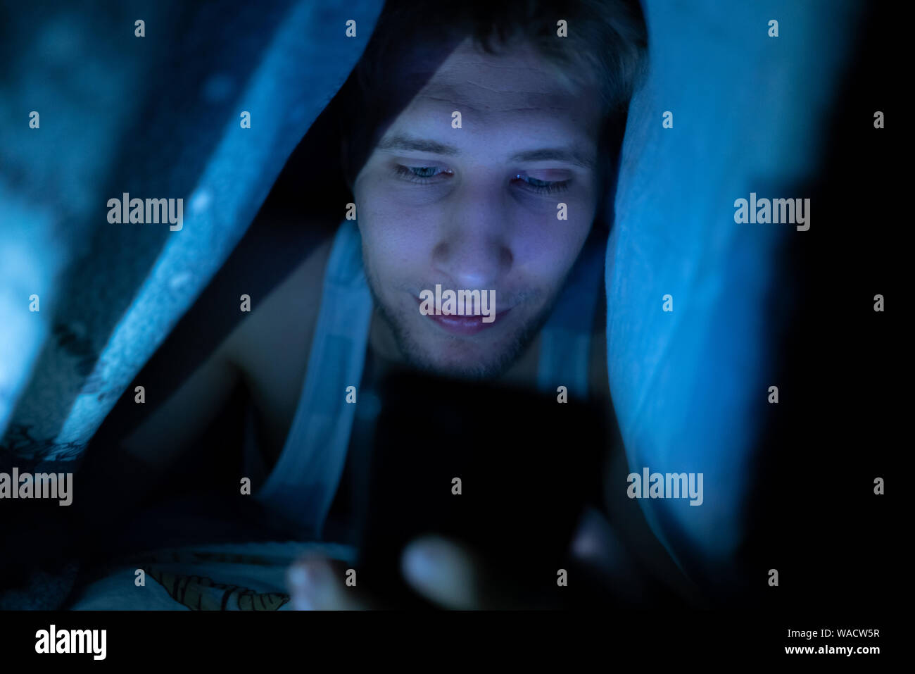 young boy using a smartphone lying in bed under a blanket at night ...