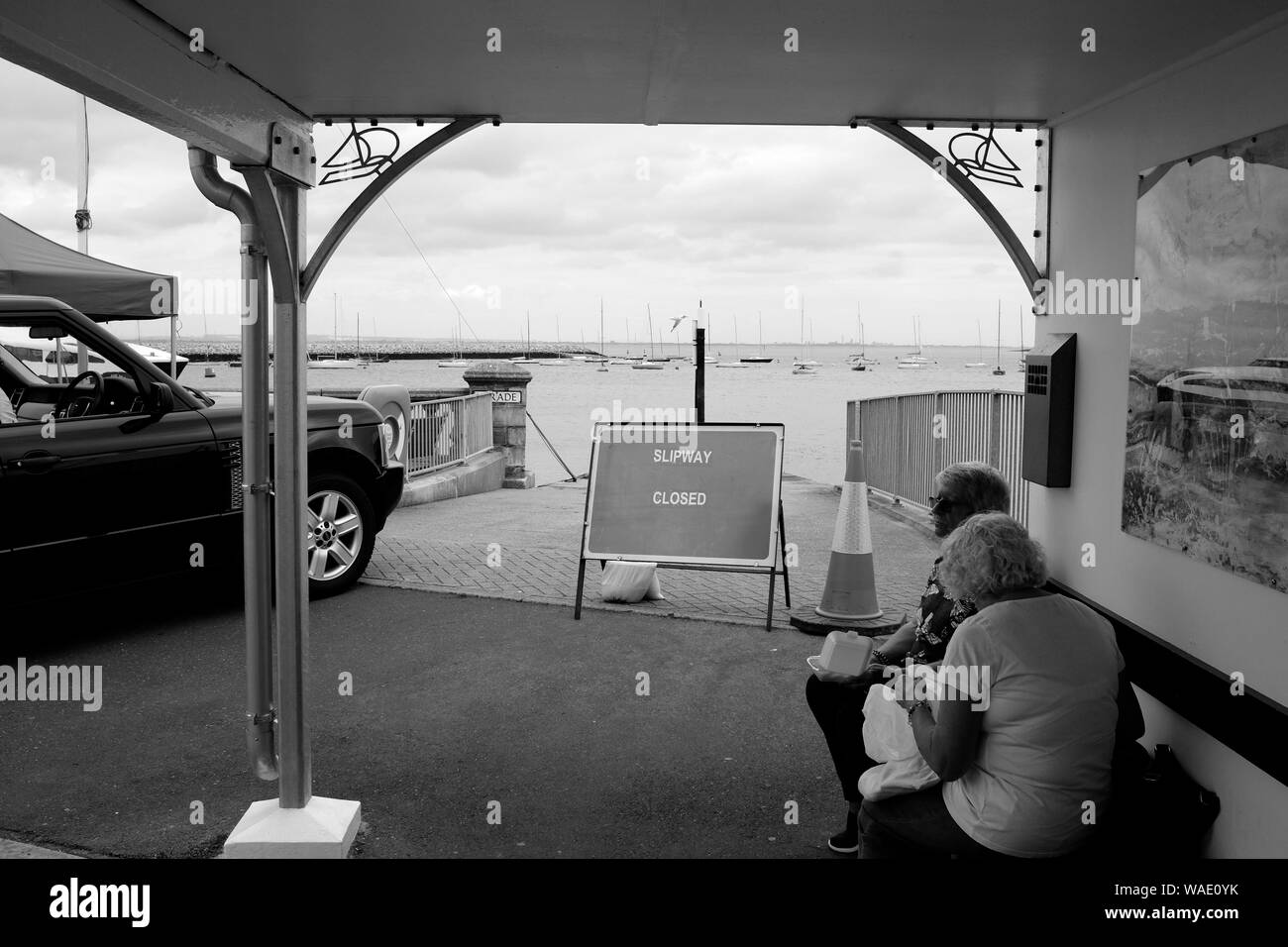 Two women sitting on a bench eating take-away food by the slipway on ...