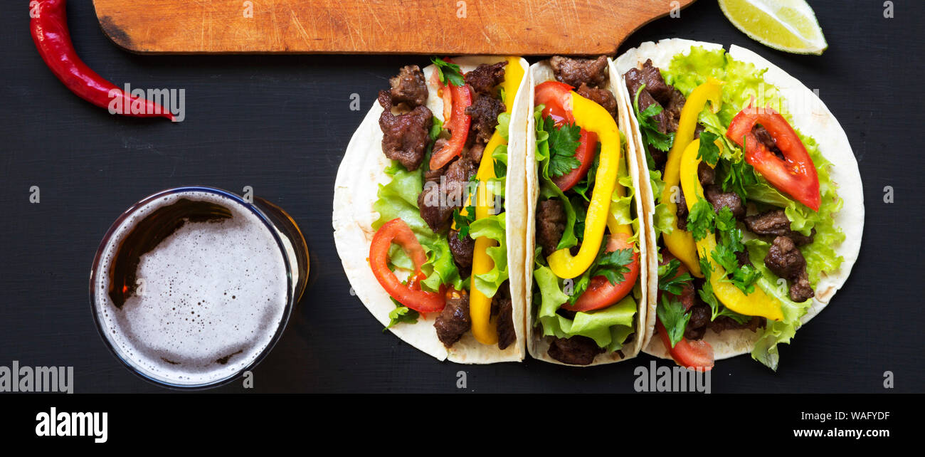 Tacos with beef and vegetables, beer and lime on a black surface, top ...