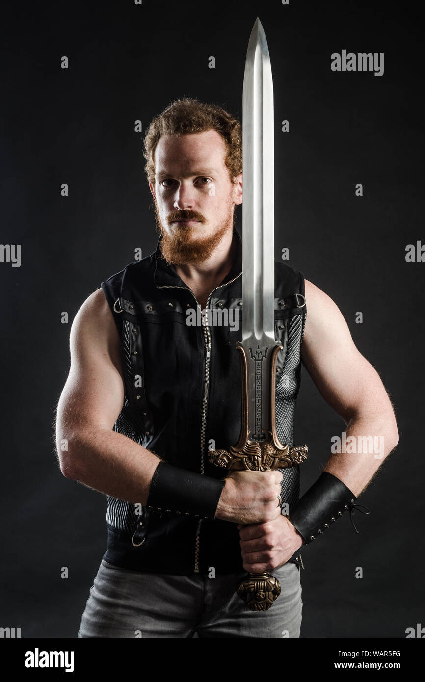 Portrait of a redhead young man with a sword and a knight coat on black ...