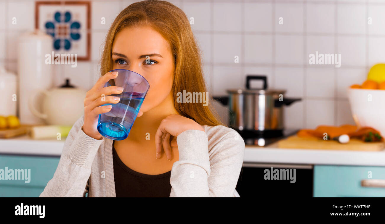 Blond woman drinking water in glass while sitting in kitchen in home ...