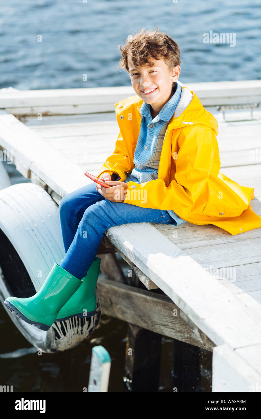 Beaming boy sitting near river wearing raincoat Stock Photo - Alamy